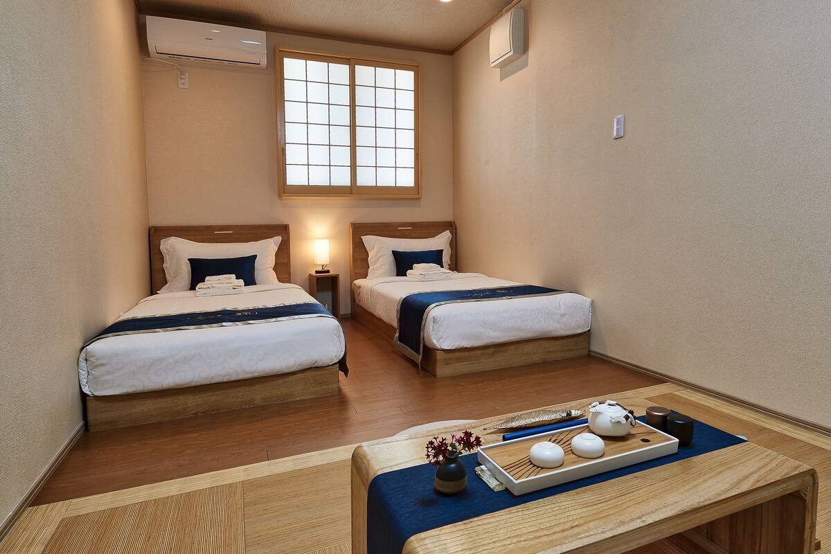 Quarto de hotel com duas camas, piso de madeira, uma mesa pequena com um tabuleiro de objetos e uma janela com persianas.