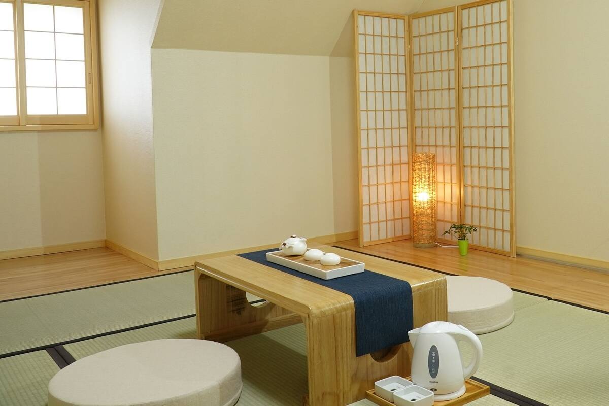 Um quarto japonês tradicional com tatames, uma mesa de madeira baixa coberta com um pano azul, uma chaleira branca e um pequeno conjunto de chá.