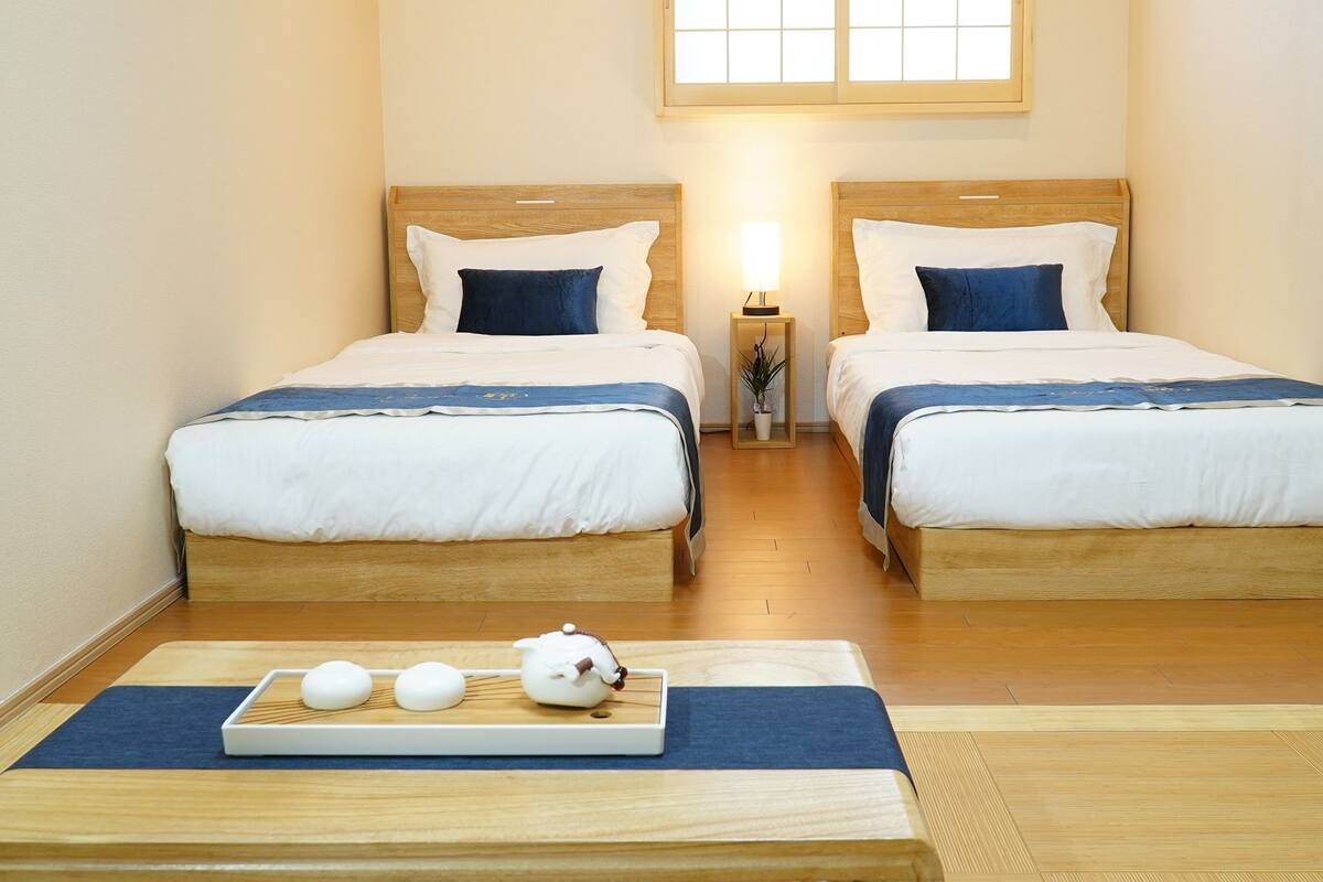 Quarto de hotel com duas camas, uma mesinha com chaleira e xícaras, e uma janela com luz natural.