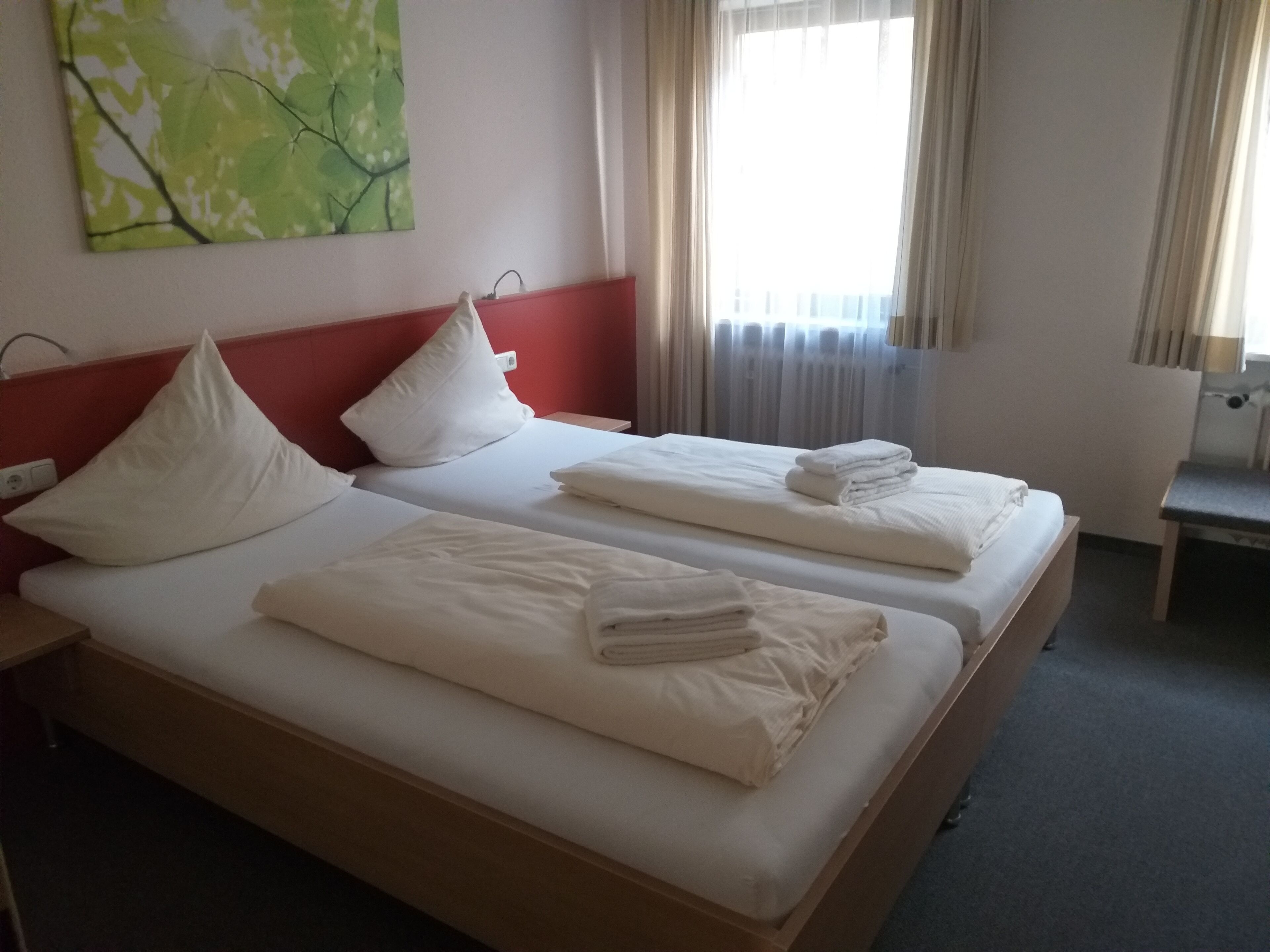Quarto de hotel com duas camas, roupa de cama branca, parede de destaque vermelha, pintura abstrata verde e uma janela com cortinas.