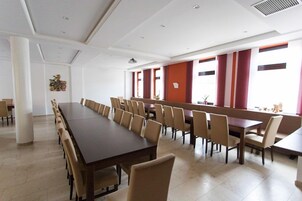 Uma sala de conferências com uma mesa comprida e várias cadeiras, janelas amplas com cortinas vermelhas e teto branco com iluminação embutida.