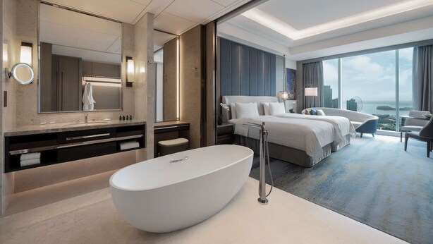 Quarto de hotel moderno com uma banheira oval grande, cama de casal, área de estar com vista para a cidade e banheiro separado com toucador e espelho.