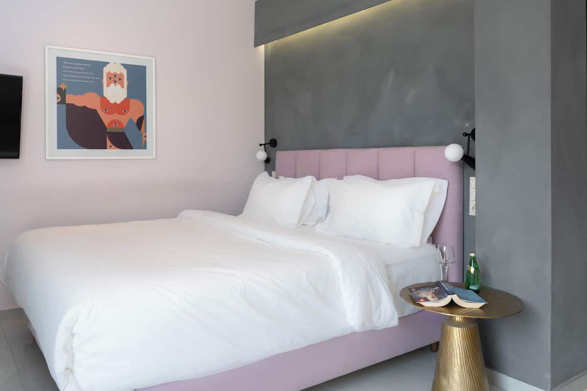 Um quarto moderno com uma cama, uma mesa de cabeceira com um livro e uma garrafa, uma televisão fixada na parede e uma obra de arte emoldurada.