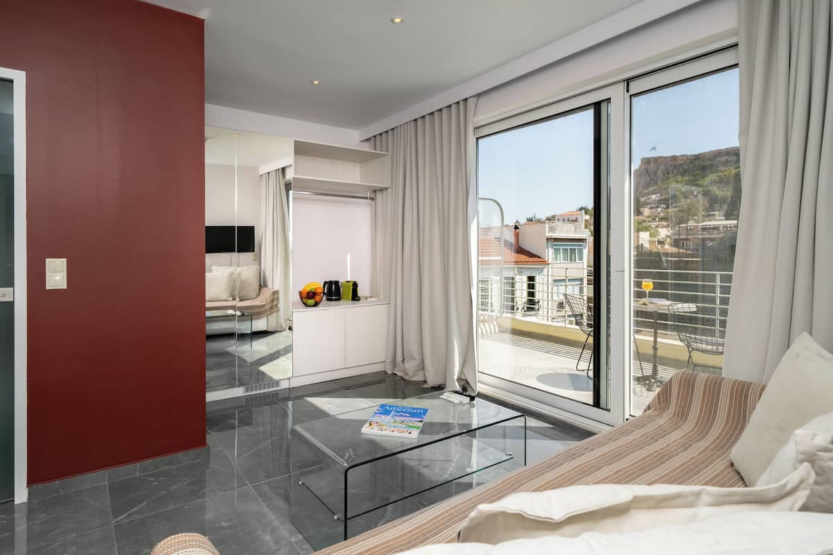 Quarto de hotel moderno com mesa de centro de vidro, sofá, televisão e varanda com vista para a cidade.