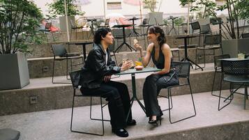 Duas pessoas sentadas à mesa com cadeiras e um copo de bebida, ao ar livre, com cadeiras e guarda-sóis ao fundo.