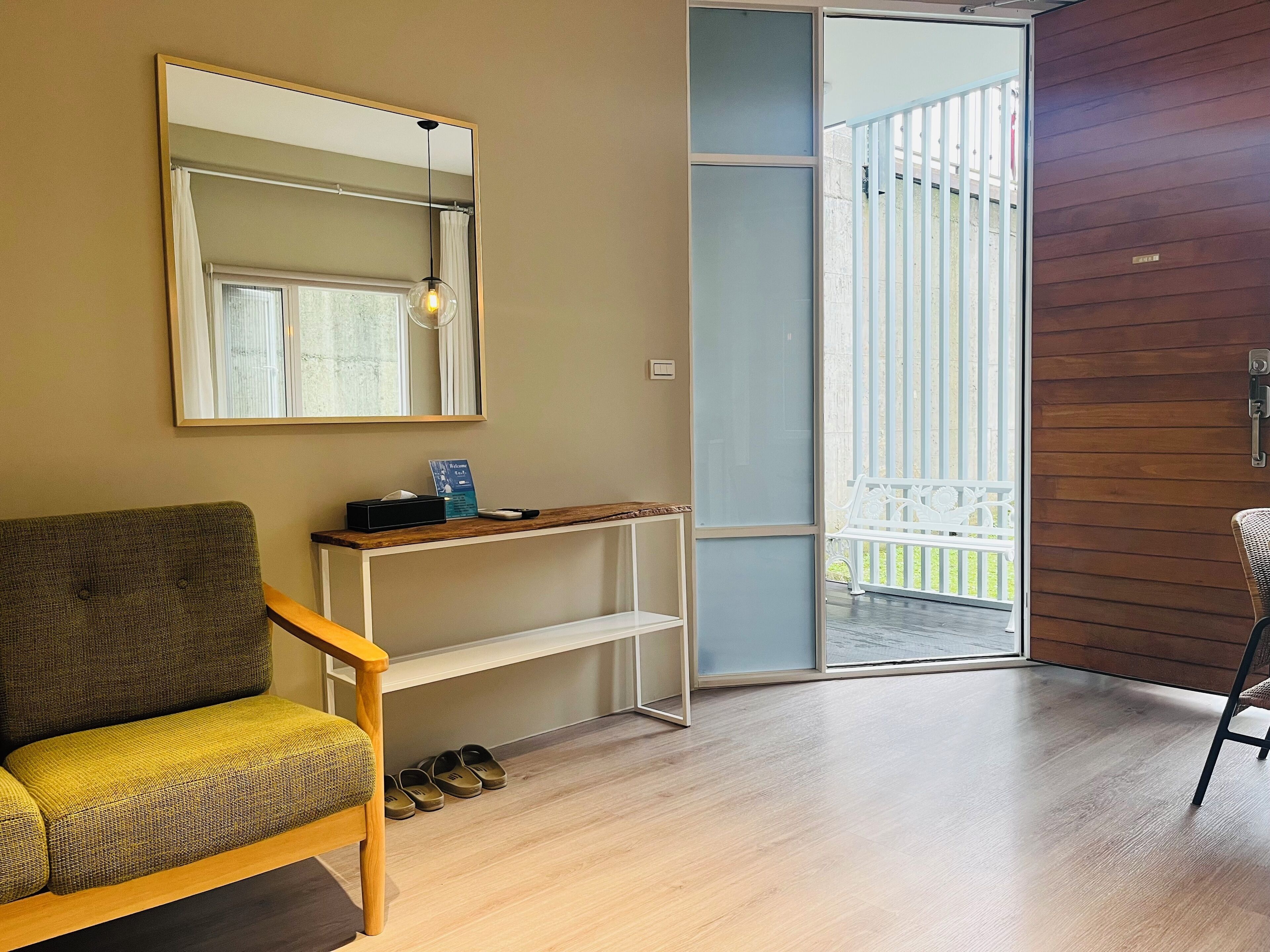 Quarto com piso de madeira, sofá, mesa de console com espelho, um banco e porta de vidro deslizante que dá para área externa com corrimão branco.