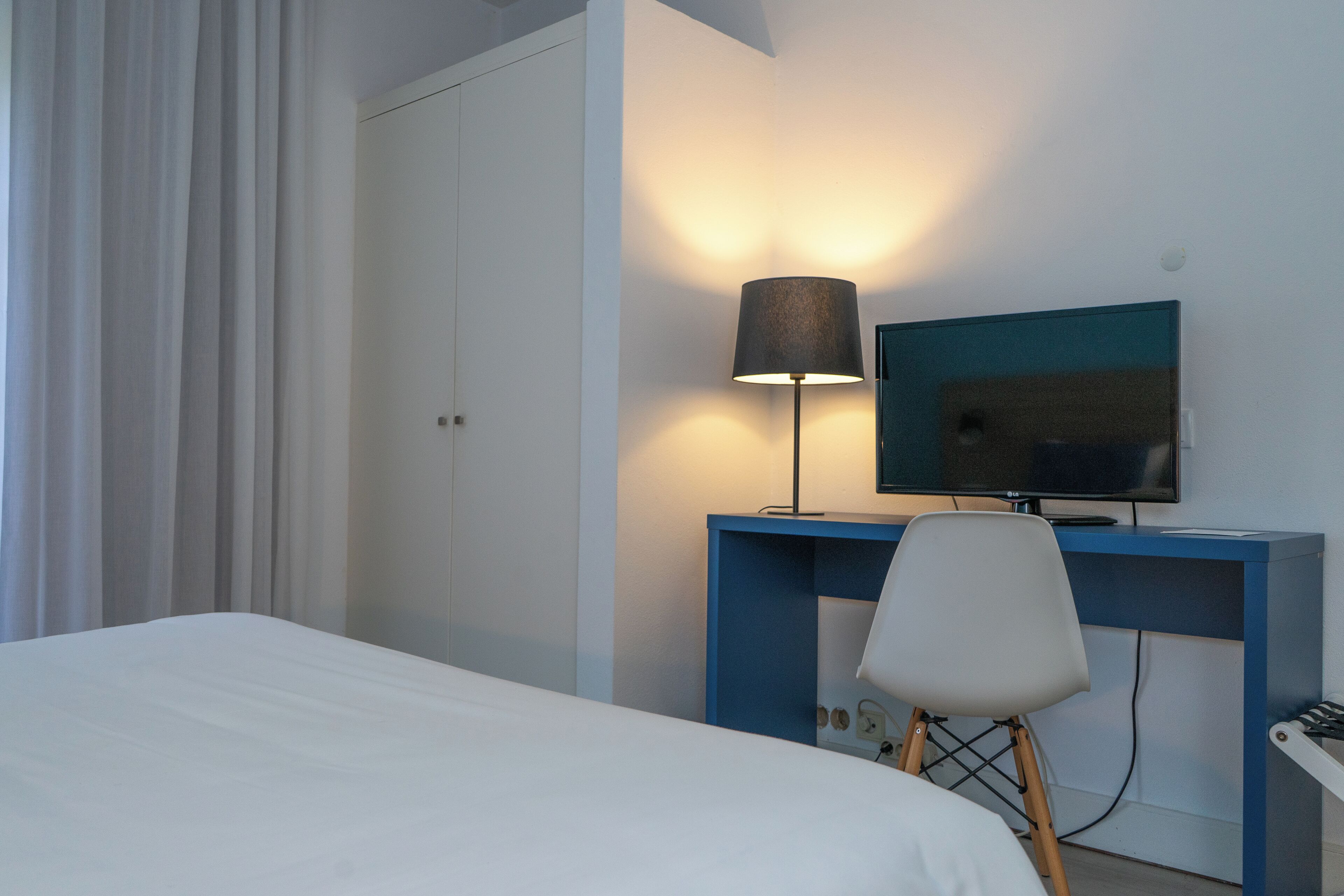 Quarto de hotel com uma cama, uma escrivaninha com luminária, uma cadeira e uma televisão.