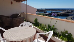 Varanda com mesa redonda e cadeiras brancas, vista para uma marina com barcos e céu azul límpido.