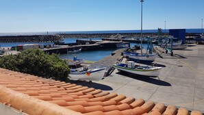 Uma marina costeira com vários barcos atracados, céu claro e um molhe rochoso ao fundo.