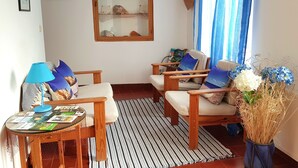 Uma sala de estar com duas poltronas de madeira, uma mesa de centro de vidro, um tapete listrado e um vaso com plantas secas.