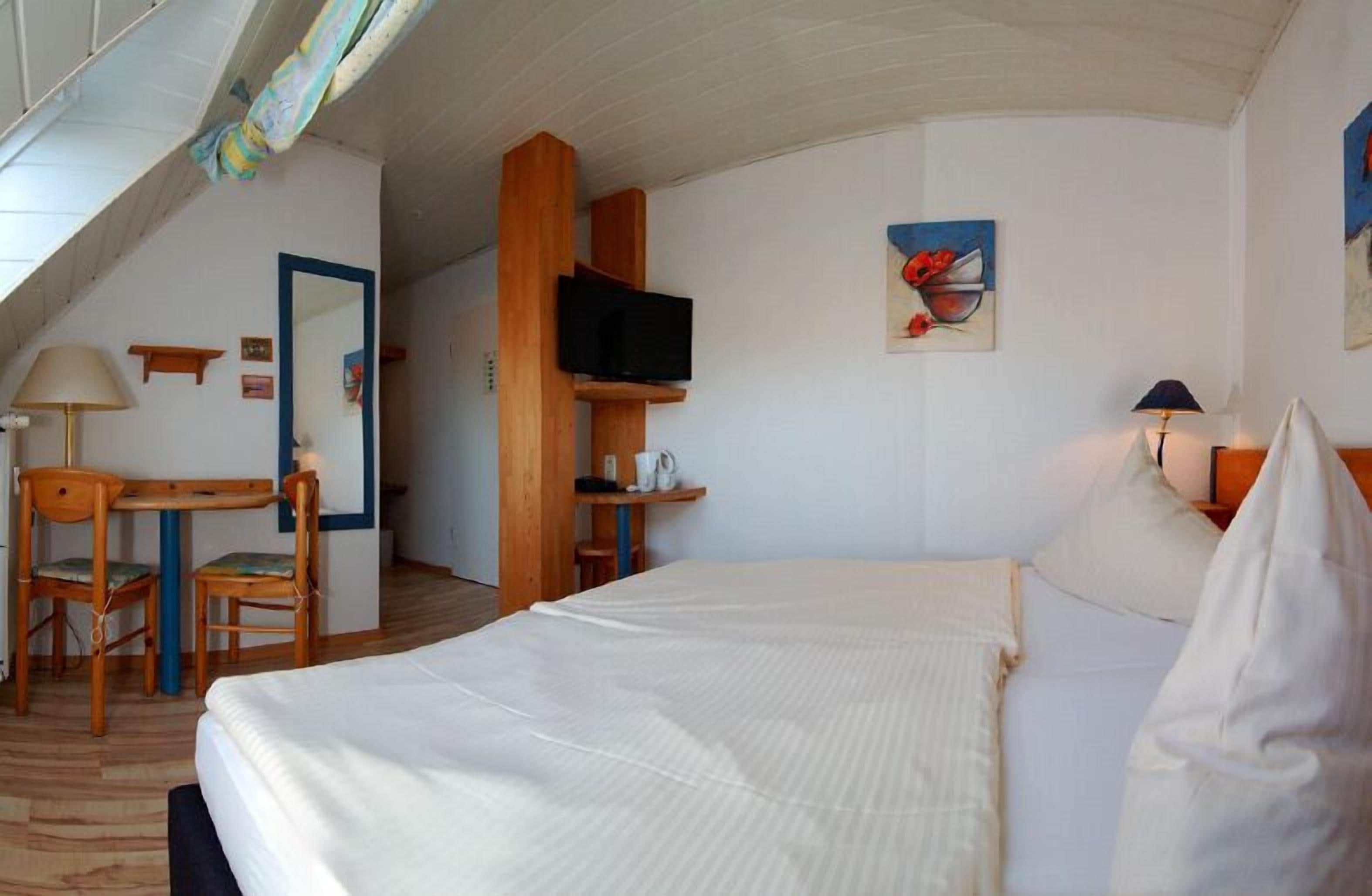 Quarto de hotel com uma cama, uma mesa com cadeira, uma televisão fixada numa prateleira de madeira e obras de arte nas paredes.
