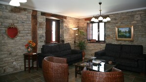 Uma sala de estar com paredes de pedra, sofás escuros, cadeiras de vime, uma mesa de centro de vidro e um quadro na parede.