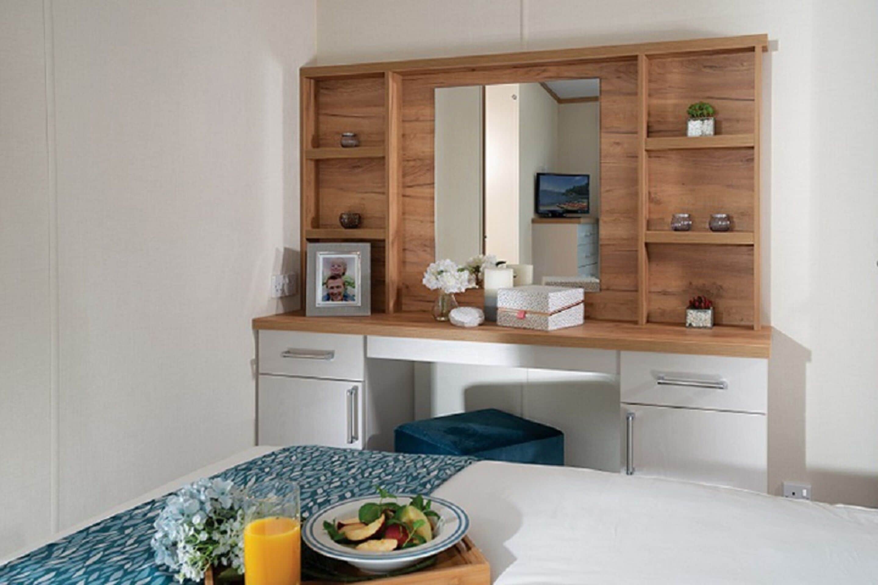 Quarto com toucador de madeira, espelho, cama com travesseiro azul, mesa de cabeceira com uma tigela de fruta e um copo de sumo de laranja, e uma pequena planta no toucador.
