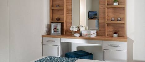 Quarto com toucador de madeira, espelho, cama com travesseiro azul, mesa de cabeceira com uma tigela de fruta e um copo de sumo de laranja, e uma pequena planta no toucador.