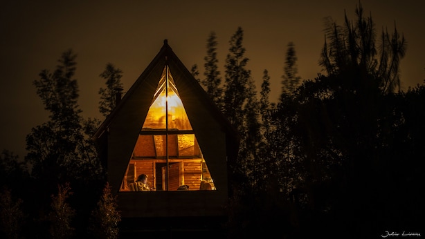 Uma cabana com telhado triangular e uma janela grande na frente, rodeada por árvores.