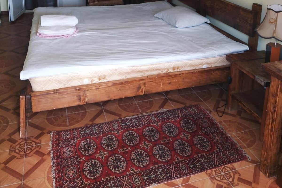 Quarto com uma cama grande, cabeceira de madeira, televisão fixada na parede, guarda-roupa de madeira, uma cadeira, tapete estampado e uma porta que dá para o banheiro.
