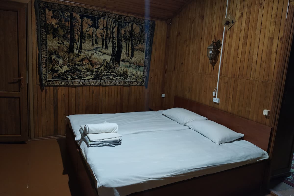 Quarto com paredes de madeira, um grande tapeçaria com tema de floresta, cama bem arrumada com lençóis brancos e uma porta de madeira.
