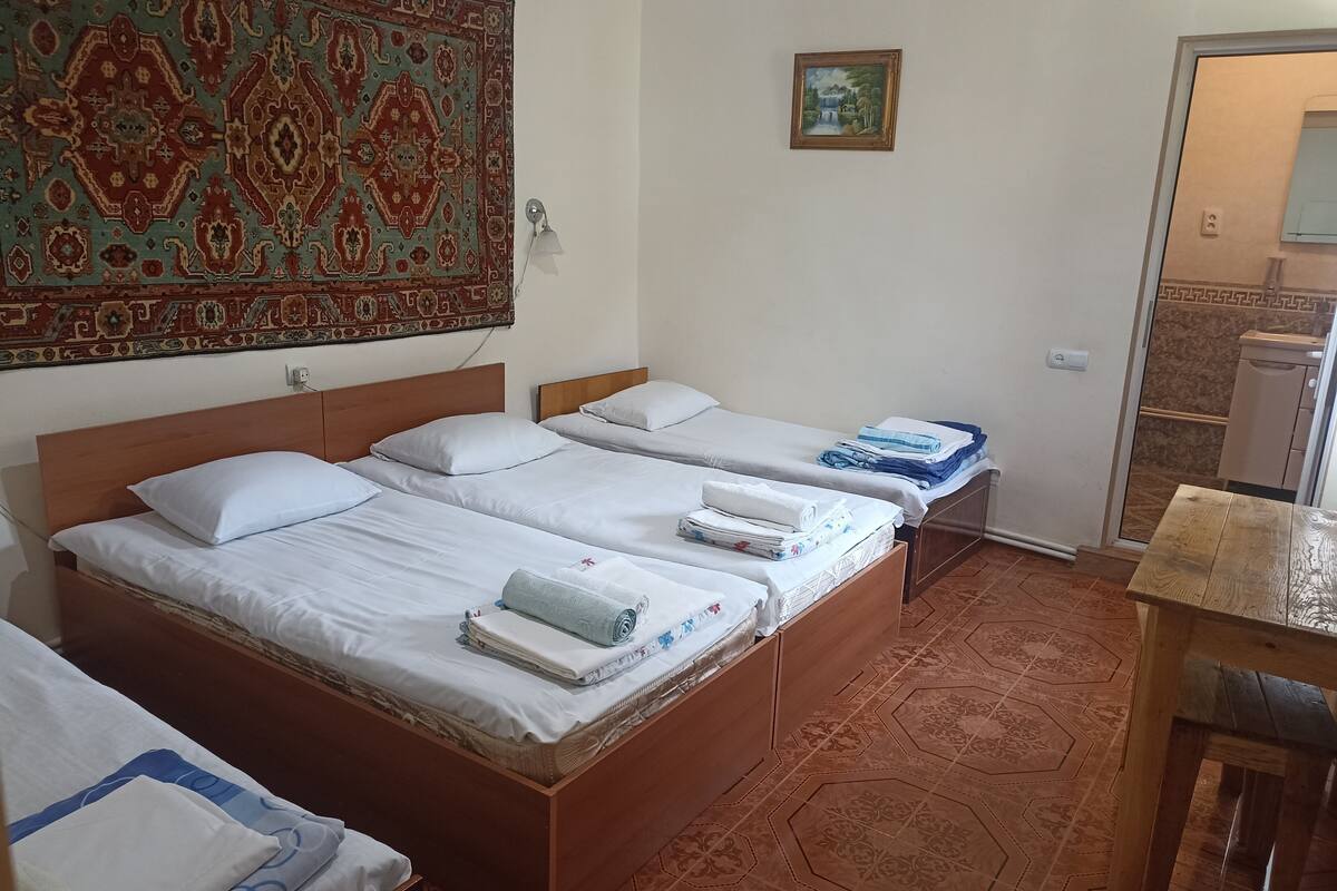 Quarto de hotel com quatro camas de solteiro, cabeceira de madeira, tapete grande com padrão, quadro emoldurado na parede e mesa de madeira.
