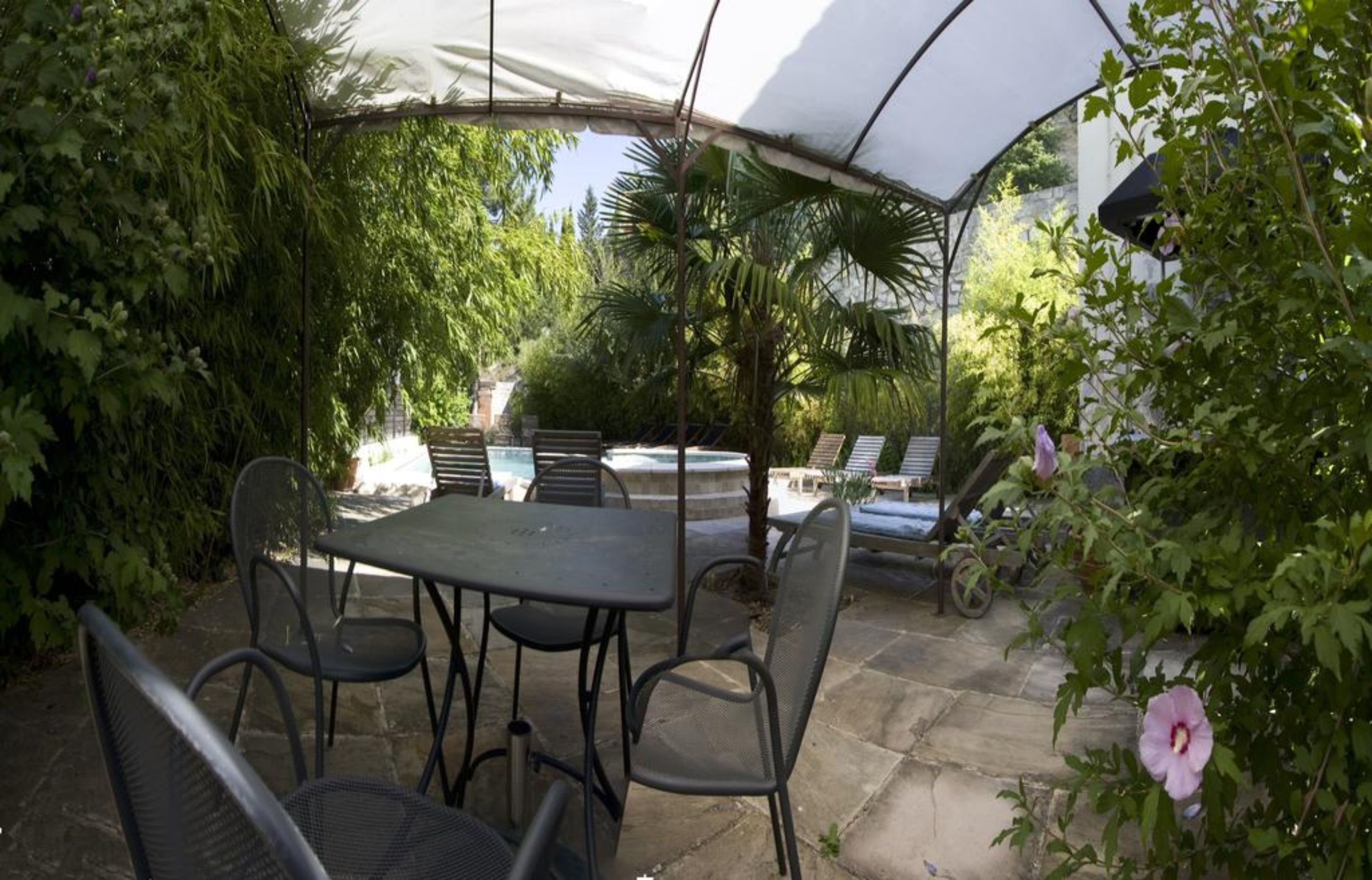 Um pátio externo com mesa e cadeiras, rodeado por vegetação exuberante e uma área de piscina.