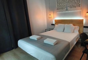 Quarto de hotel com uma cama grande, cabeceira, duas mesinhas de cabeceira, uma cadeira e uma obra de arte pendurada na parede.