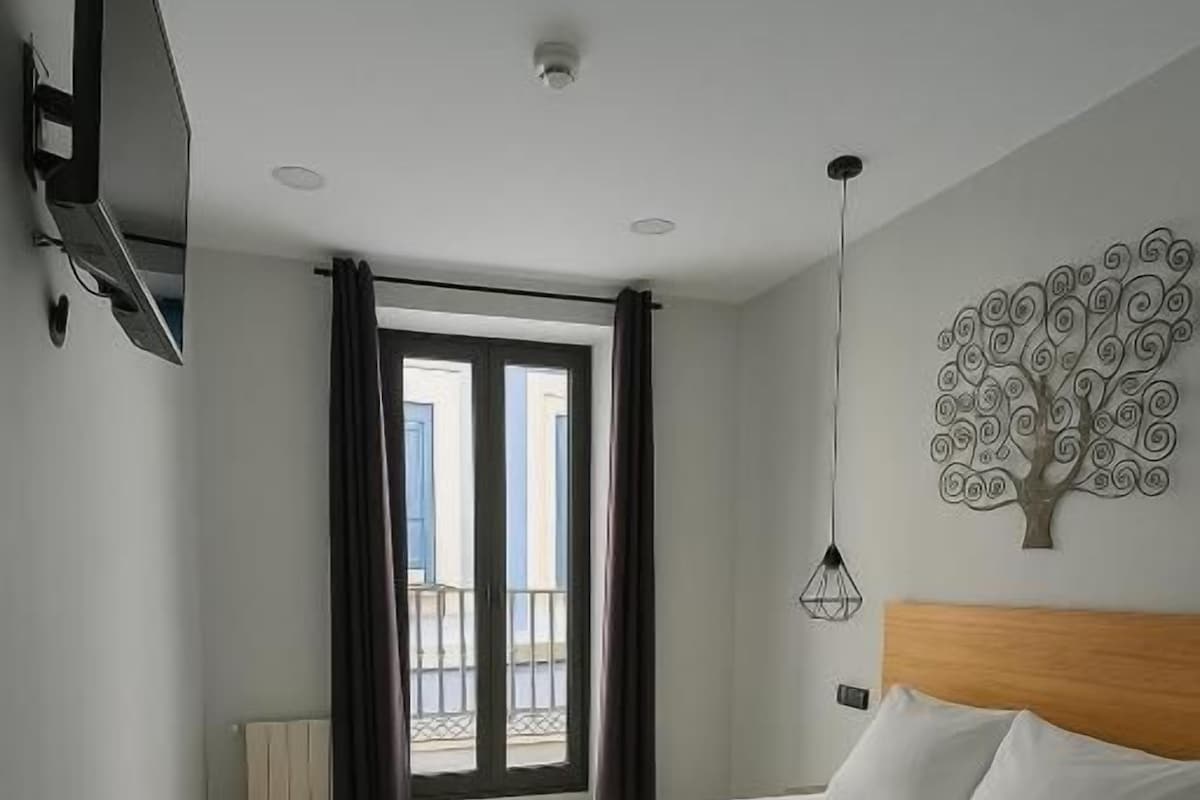 Quarto de hotel moderno com uma cama grande, televisão de tela plana, varanda com cortinas e um detalhe decorativo na parede.