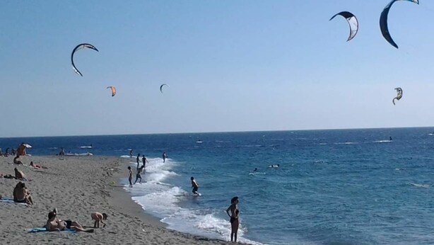 Uma praia com pessoas se banhando ao sol e vários pipas voando no céu.