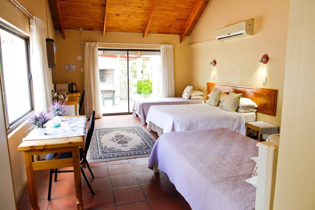 Quarto com duas camas, uma mesa de madeira com uma planta, uma cadeira, uma mesinha com chaleira e vista para o verde lá fora.