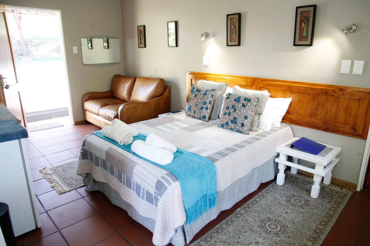 Quarto bem arrumado com uma cama, uma mesinha de cabeceira branca, uma poltrona de couro marrom e uma porta que dá para o exterior.