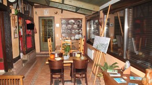 Uma área de refeições com uma mesa e cadeiras de madeira, uma pintura em cavalete e uma estante com louças.