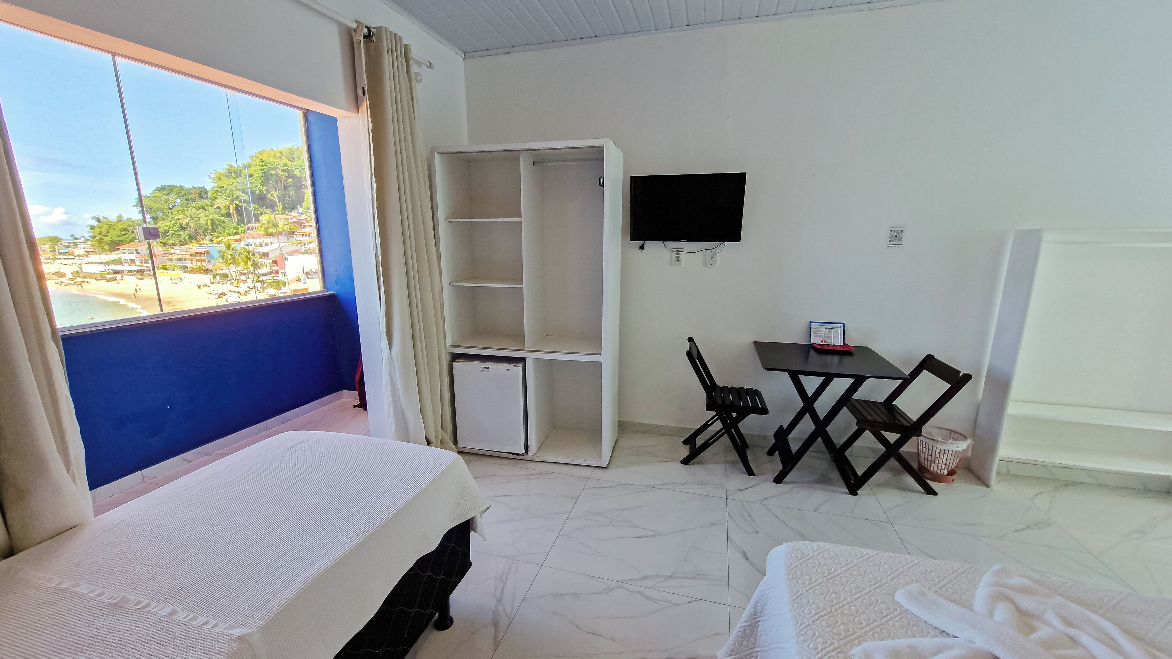 Quarto de hotel com cama, escrivaninha com cadeira, televisão e vista para a praia.