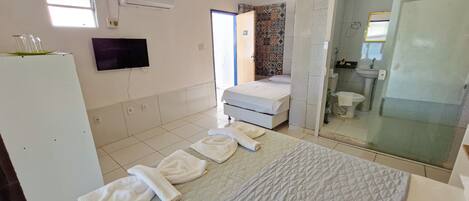 Quarto de hotel com cama, televisão, ar condicionado, banheiro com vaso sanitário e pia, e chuveiro.