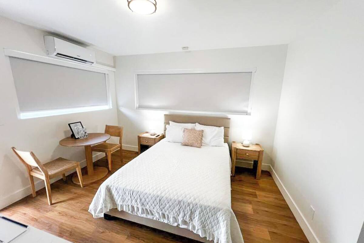 Quarto com cama, mesas de cabeceira, uma cadeira, escrivaninha e janela.