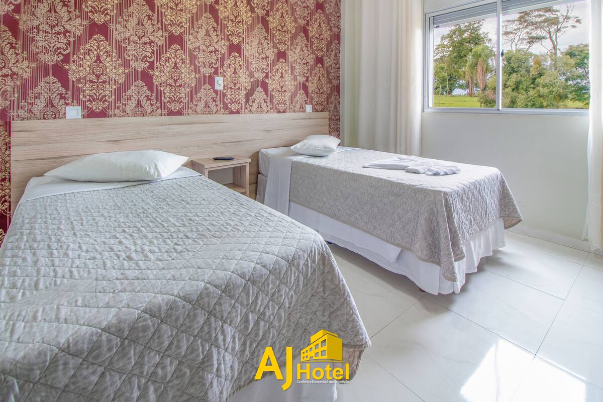 Quarto de hotel com duas camas, cabeceira de madeira, papel de parede estampado e janela com vista para árvores.