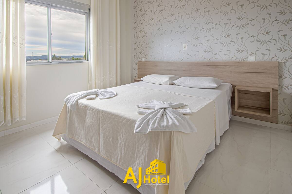 Quarto de hotel com uma cama grande, cabeceira de madeira e vista pela janela.