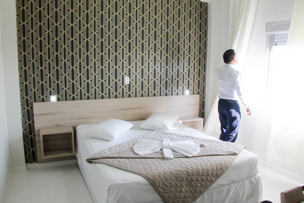 Quarto de hotel com cama bem arrumada, cabeceira com padrão geométrico e vista para o exterior através de uma janela.