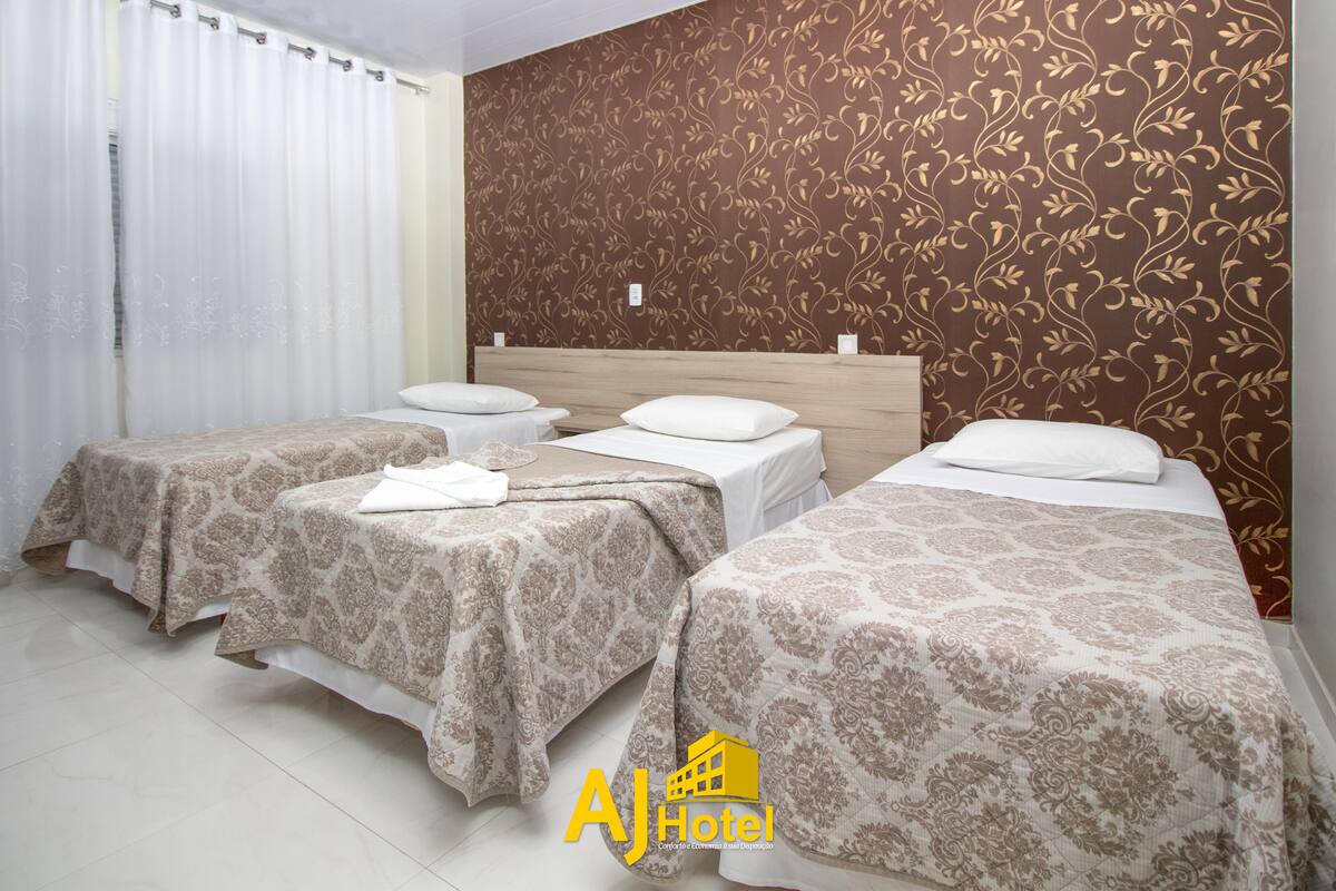 Quarto de hotel com três camas, roupa de cama branca, cabeceira de madeira e papel de parede estampado.