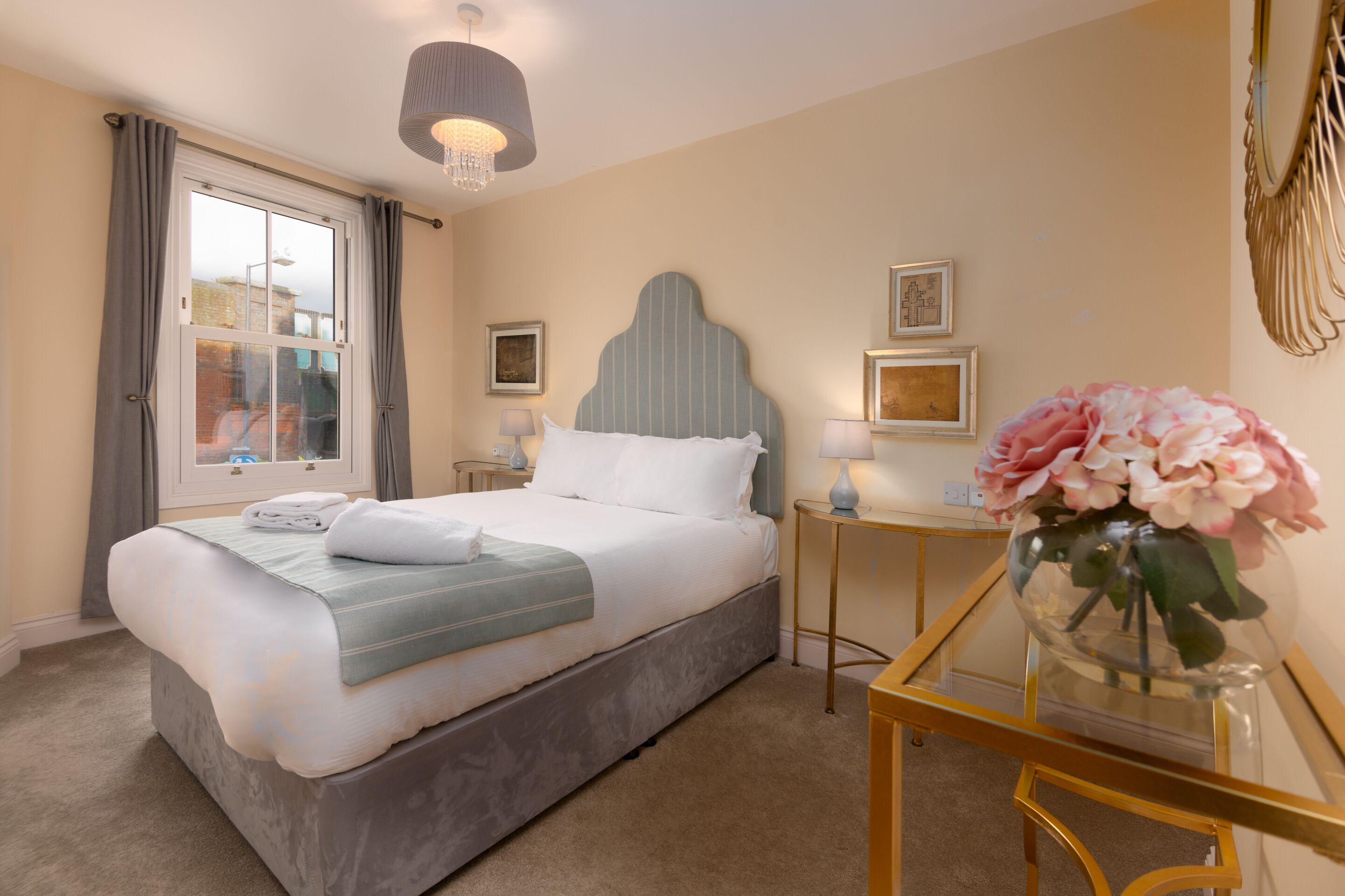 Quarto de hotel com cama, mesas de cabeceira, escrivaninha, cadeira, janela com cortinas e um vaso com flores.