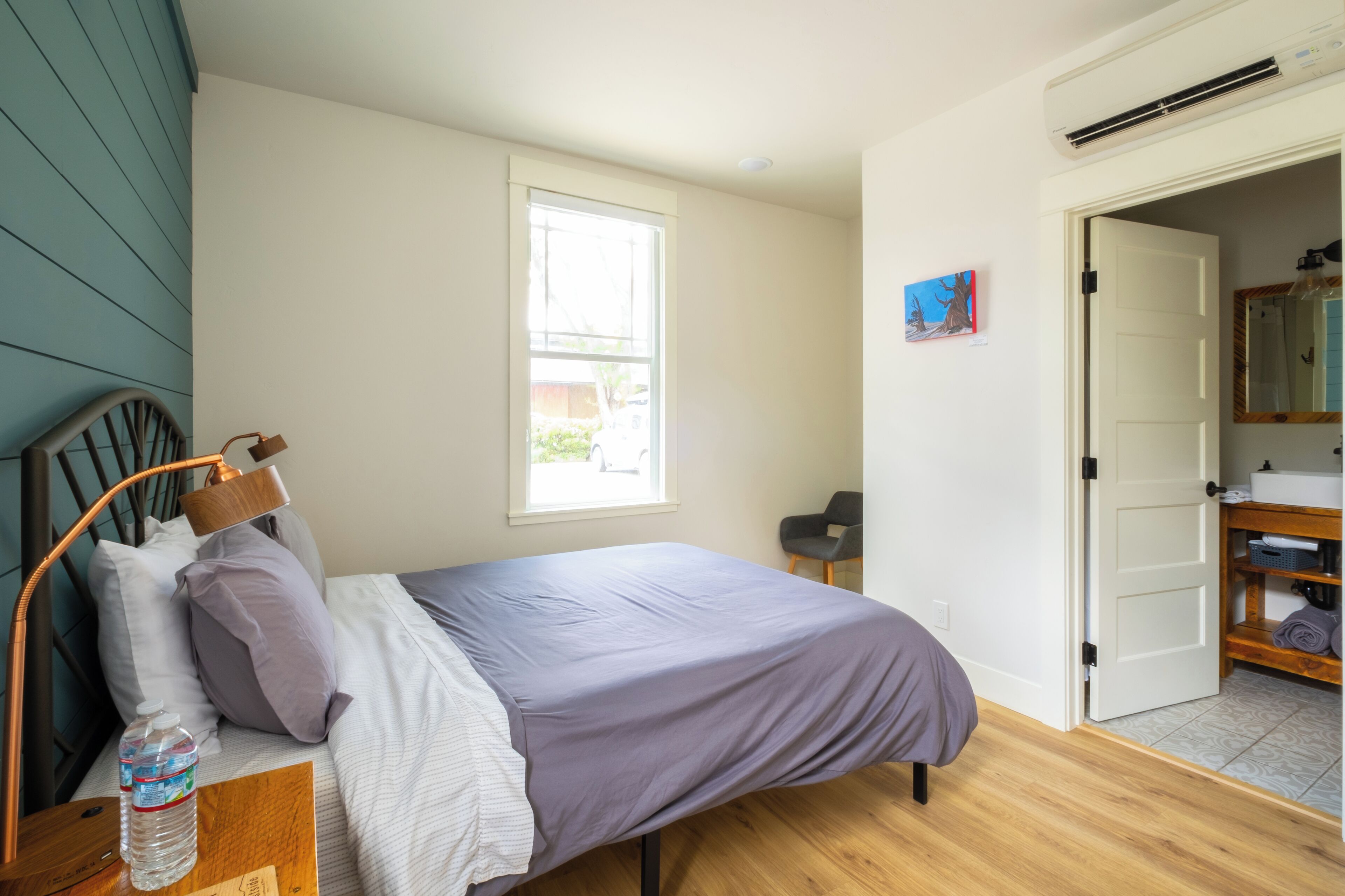 Quarto com uma cama, mesa de cabeceira, garrafa de água, janela, porta e banheiro visível pela porta.