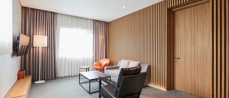 Quarto de hotel moderno com sofá, poltrona e uma pequena mesa de centro. Há uma TV fixada na parede e uma porta de madeira.