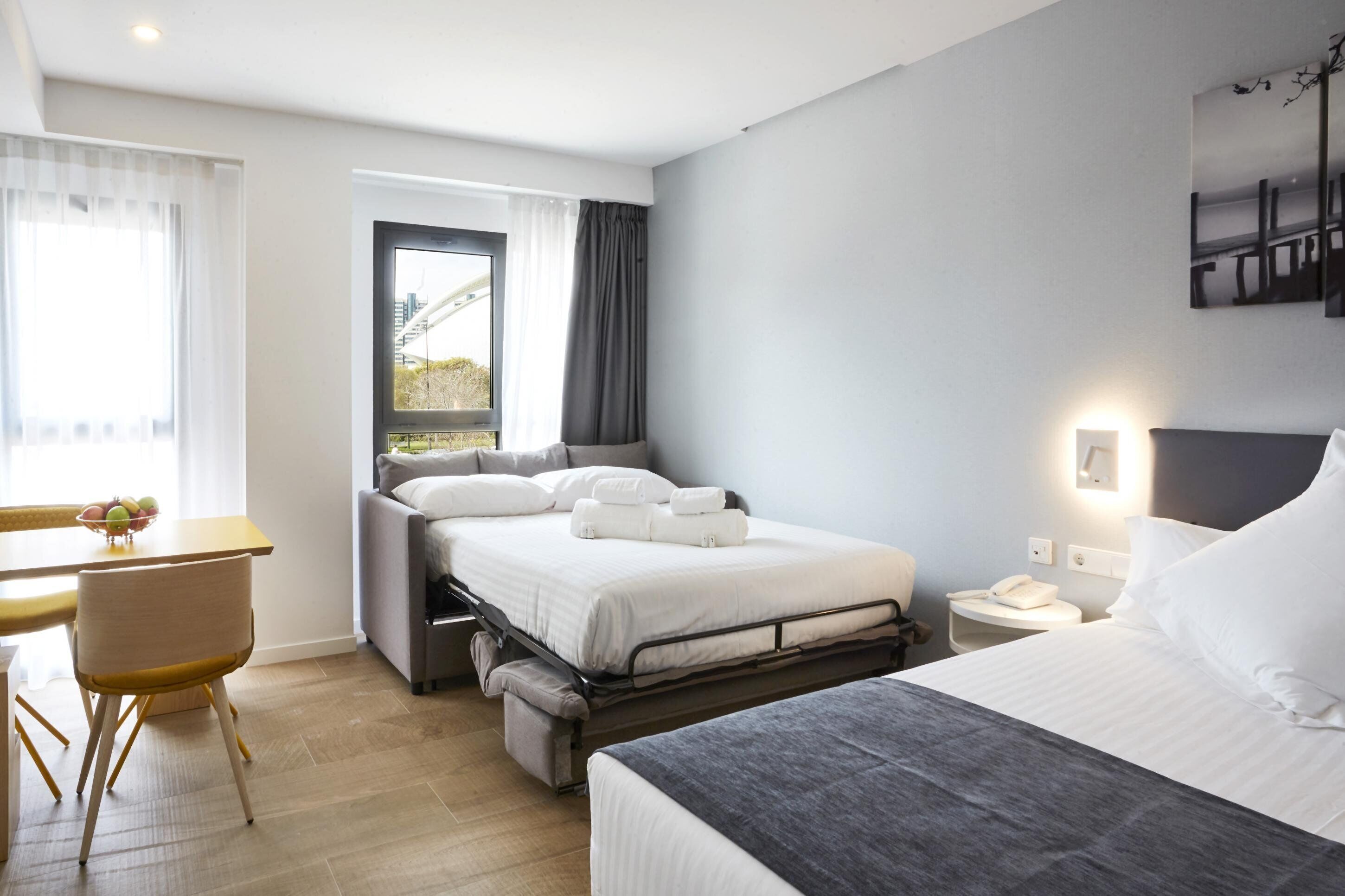 Quarto de hotel com cama, escrivaninha e cadeira. Possui janela com cortinas, mesa de cabeceira com abajur e telefone fixado na parede.