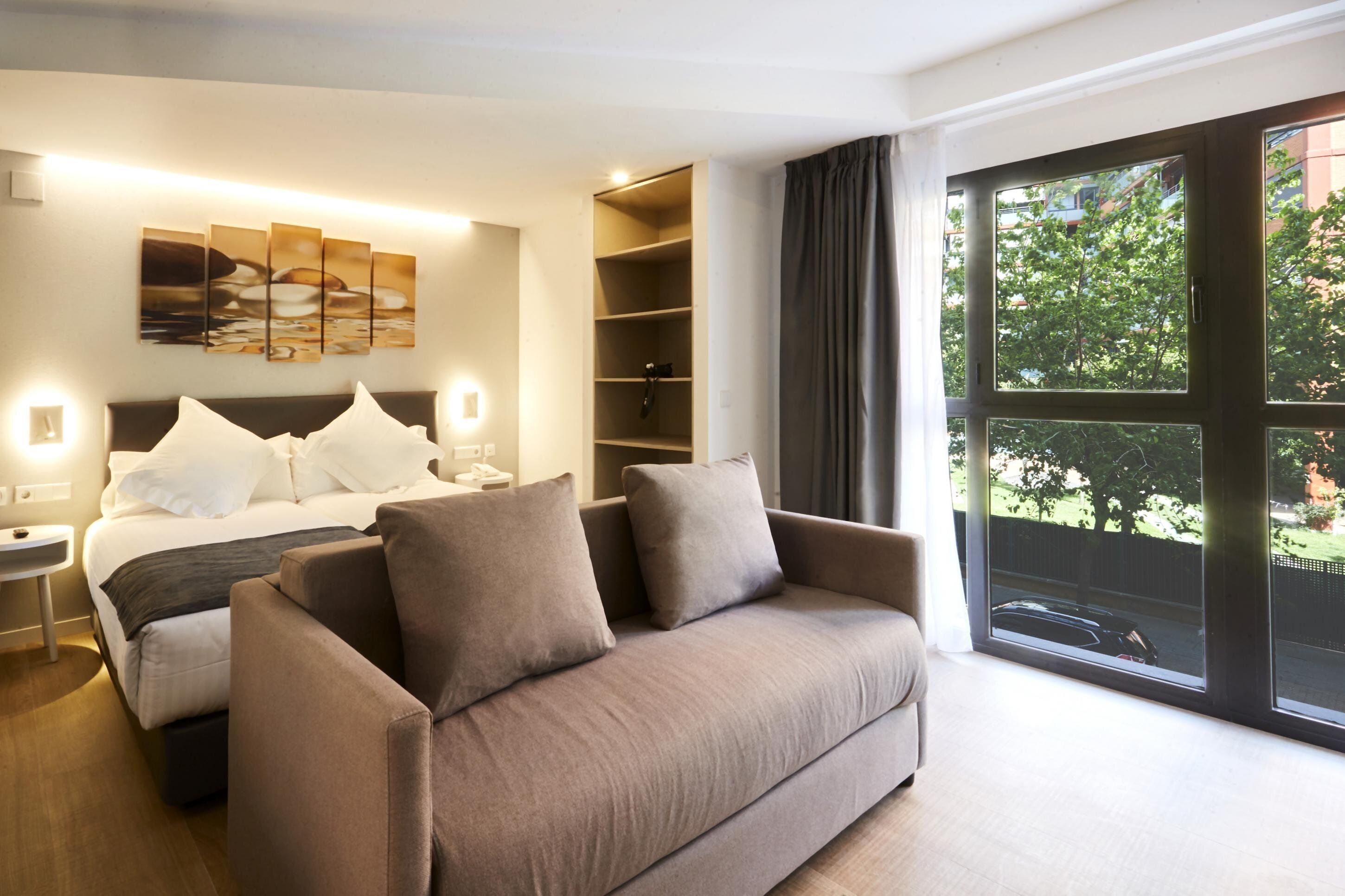 Quarto de hotel moderno com cama, sofá, uma grande janela e uma obra de arte pendurada na parede.