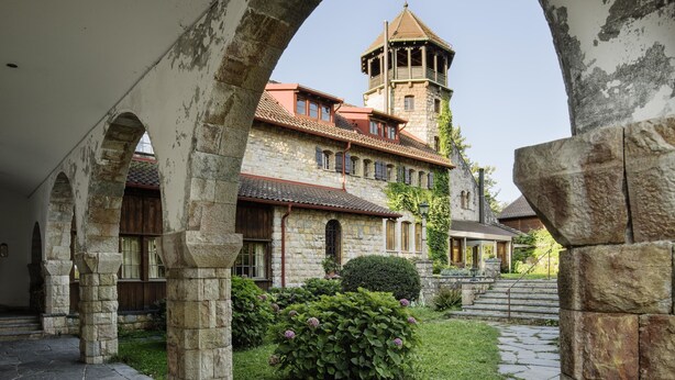 Um edifício de pedra com uma torre, passarelas em arco e um jardim bem cuidado.