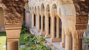 Um corredor de pedra com arcos e colunas, ladeado por um jardim florido e repleto de plantas.