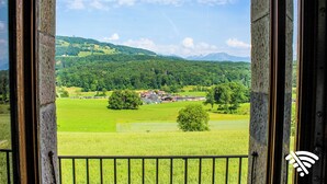 Vista de varanda com guarda-corrimão de metal, sobre paisagem verde com árvores, um pequeno edifício e colinas ao longe.