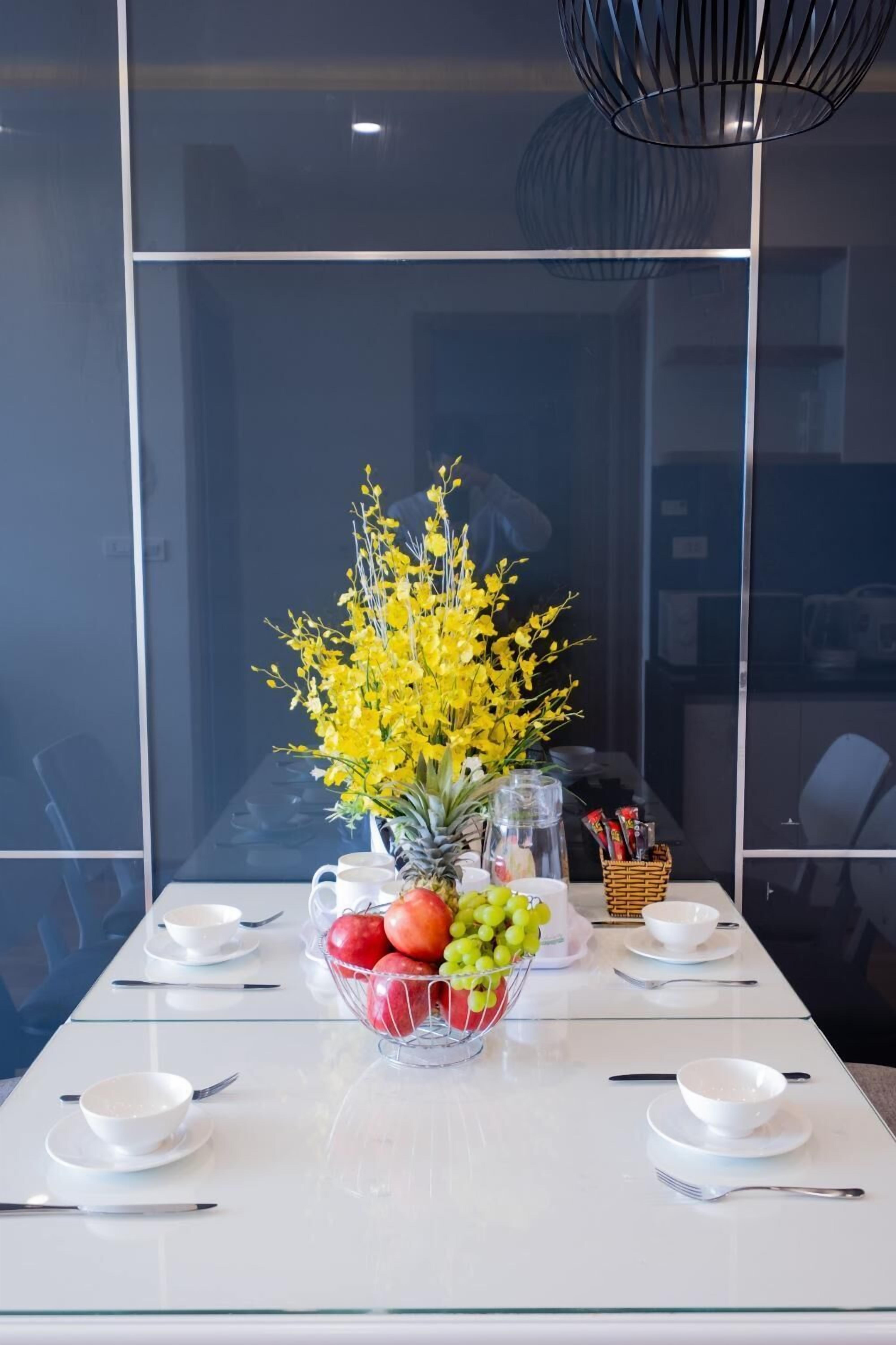 Mesa posta com pratos, tigelas e talheres brancos, acompanhada de uma fruteira e um vaso de flores amarelas.