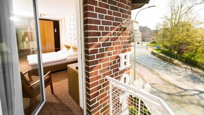 Quarto de hotel com cama, uma mesinha e uma cadeira. Tem uma varanda com vista para a rua e para as árvores.