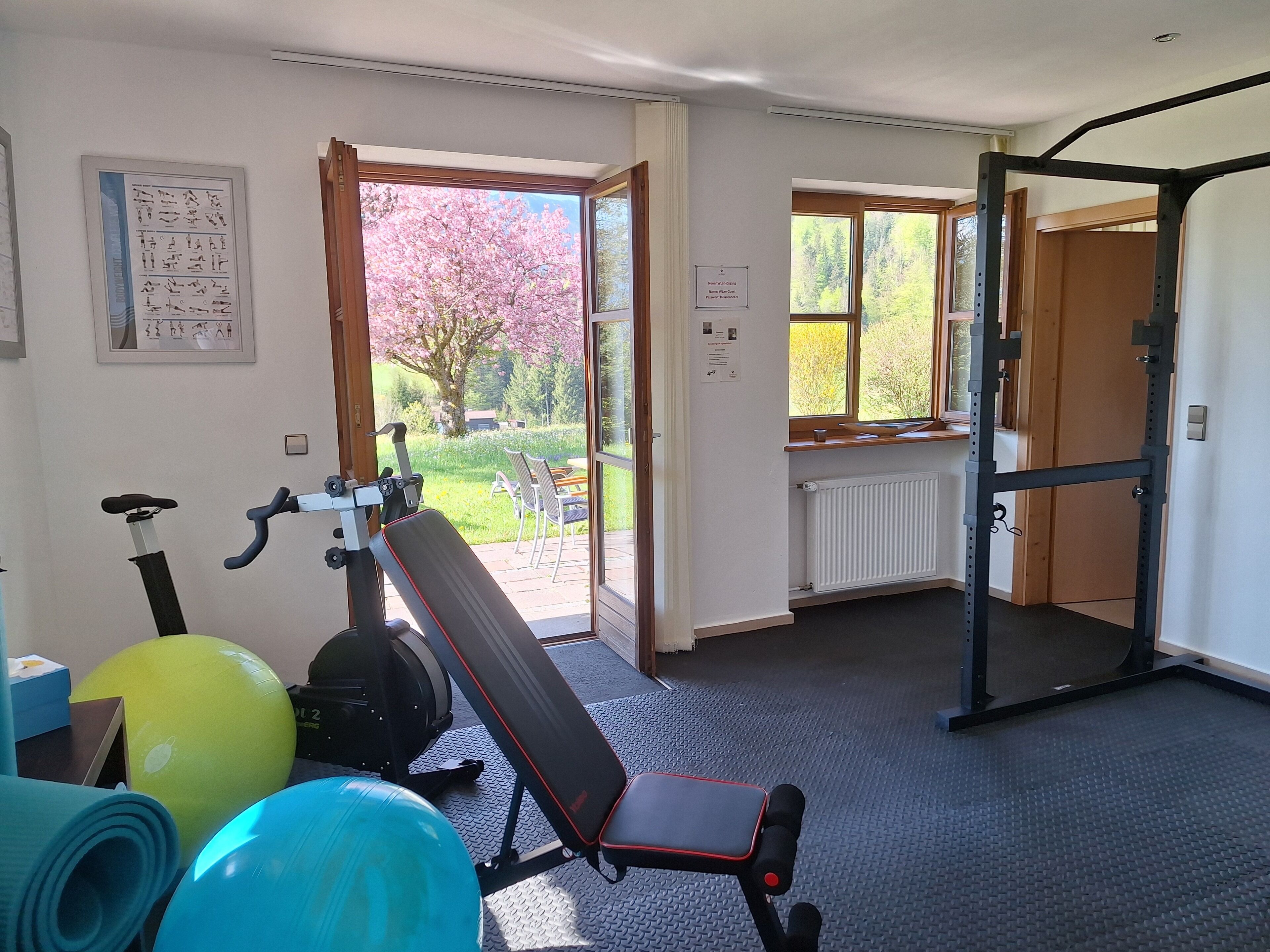 Uma academia com equipamentos de exercícios, um banco e vista para um jardim pela porta.