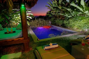 Uma área de piscina com vista para o pôr do sol, rodeada por vegetação exuberante e uma estrutura com telhado de palha.