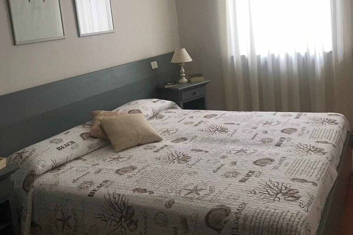 Quarto com cama, duas fotos emolduradas na parede, mesa de cabeceira com abajur e janela com cortinas.