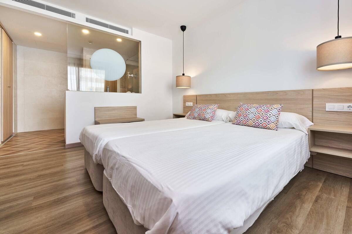 Quarto de hotel moderno com cama grande, cabeceira de madeira, mesas de cabeceira, espelho e luminária pendente.